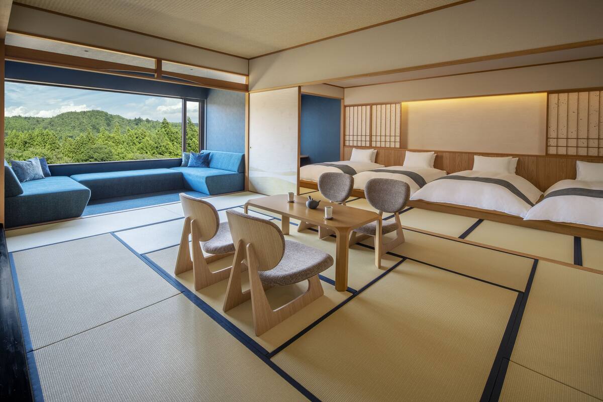 Um quarto em estilo japonês tradicional, com tatame, uma mesa baixa com cadeiras, um sofá e uma cama com cabeceira de madeira.
