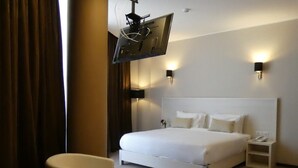 Quarto de hotel com uma cama grande, televisão presa na parede, mesa de cabeceira com telefone e uma mesa redonda com uma cadeira.