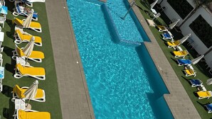 Piscina retangular com um deck ladrilhado, rodeada por espreguiçadeiras e guarda-sóis.