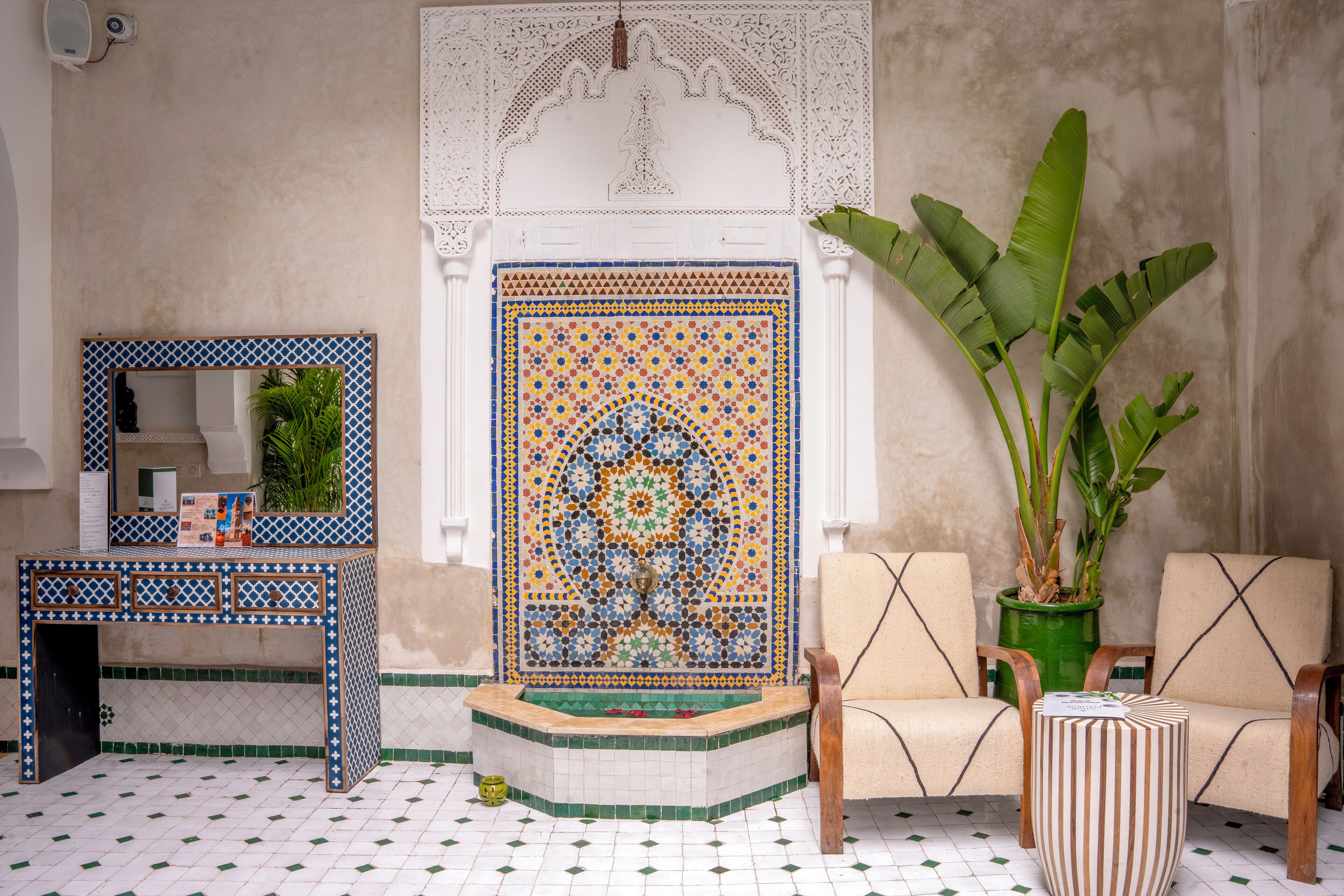Um quarto com piso de azulejos, parede decorativa com padrão geométrico, espelho, uma pequena mesa, um vaso com planta e duas cadeiras.