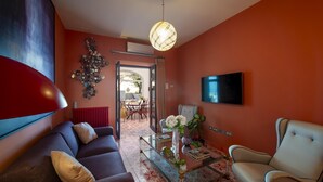 Sala de estar com sofá, poltrona, mesa de centro e objetos decorativos. Há uma televisão fixada na parede e uma porta que dá para outro cômodo.