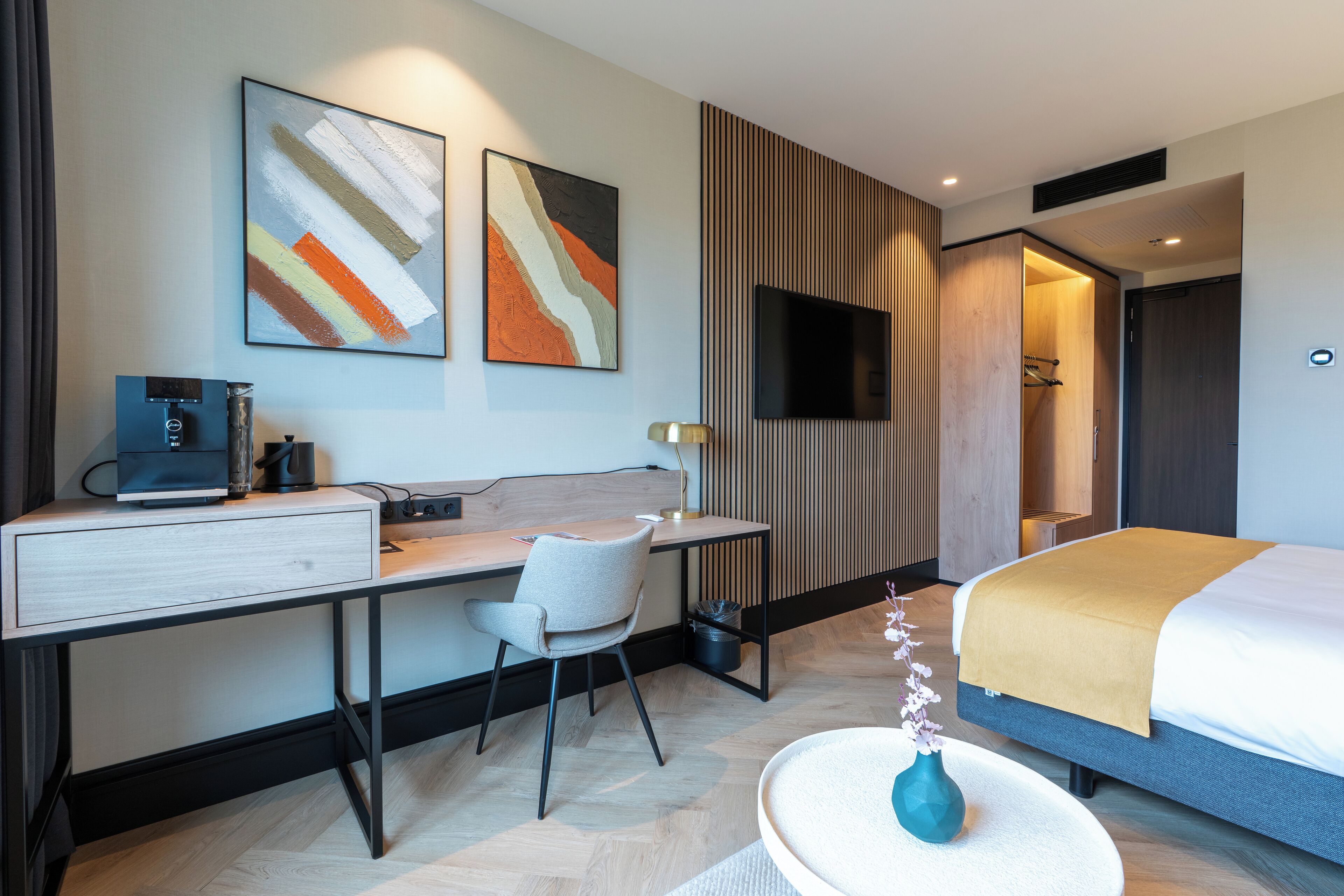 Quarto de hotel moderno com escrivaninha, cadeira, cama, TV e obra de arte na parede.