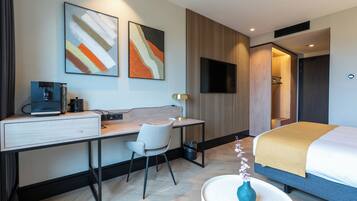 Quarto de hotel moderno com escrivaninha, cadeira, cama, TV e obra de arte na parede.