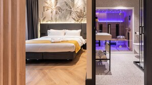Quarto de hotel moderno com uma cama grande, banheiro com paredes de vidro e área de spa com iluminação azul.