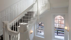Uma escadaria branca com design curvo, uma janela com vista para a rua e um candeeiro fixado na parede.