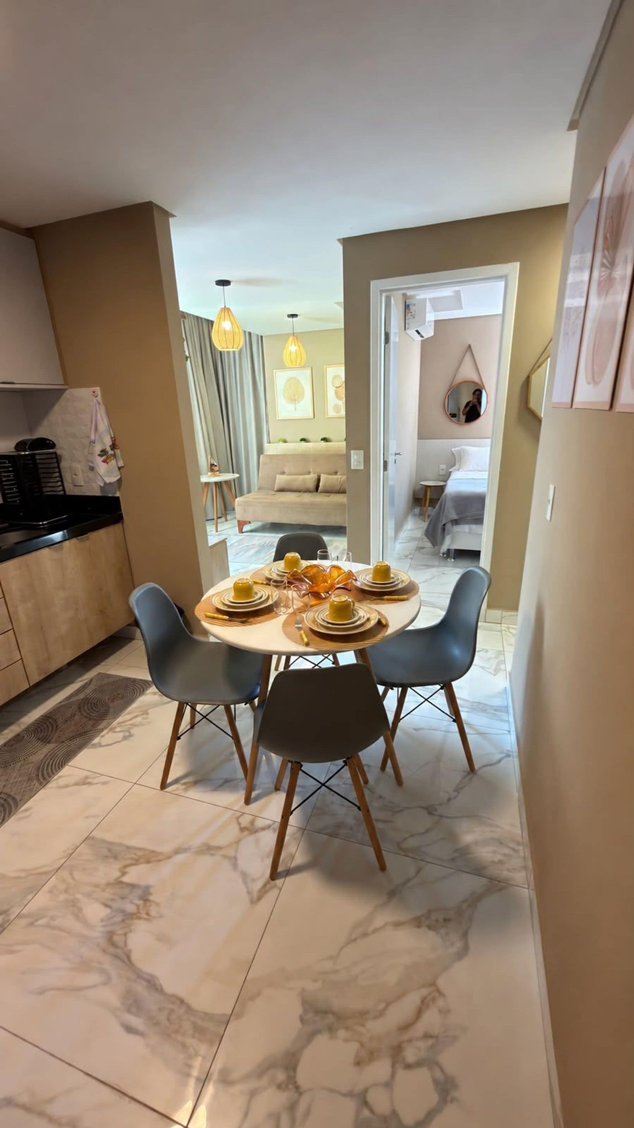 Uma sala de jantar com uma mesa redonda posta para quatro pessoas, rodeada por cadeiras azuis, num apartamento moderno onde se vê uma cozinha e uma sala de estar ao fundo.