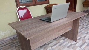 Uma escrivaninha de madeira com um laptop Lenovo, uma cadeira vermelha e uma janela com grades.