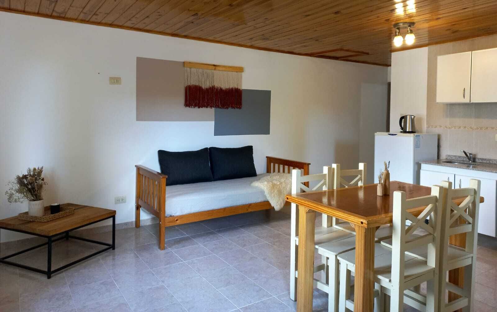 Um quarto aconchegante com um sofá de madeira, uma mesa de jantar com cadeiras, uma pequena mesa lateral e uma cozinha com armários e geladeira.
