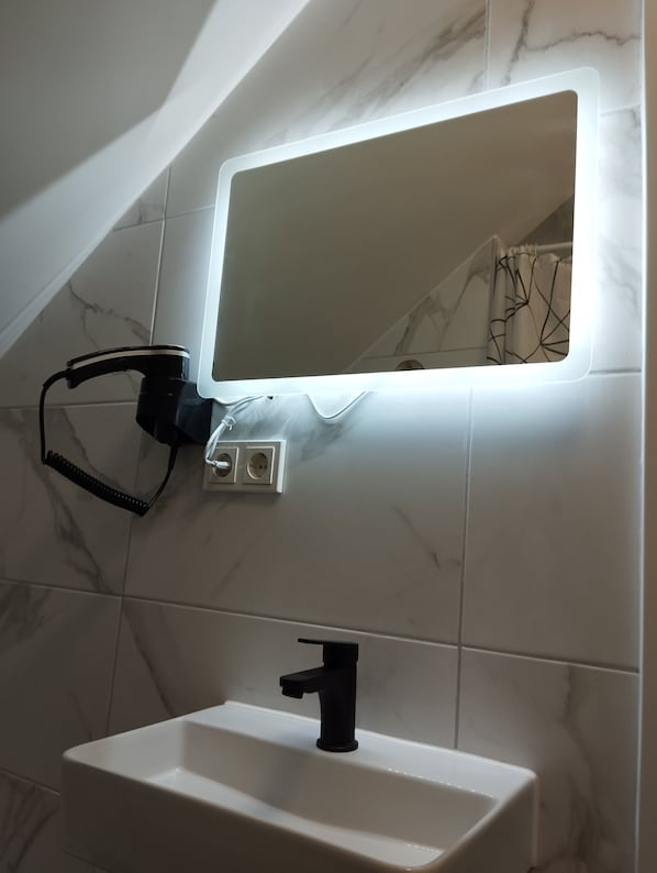 Casa de banho com uma pia branca, torneira preta, espelho com iluminação embutida e secador de cabelo na parede.