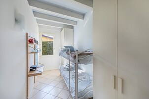 Quarto em dormitório com beliches, uma estante com livros e um objeto vermelho, e uma porta que dá para uma varanda.