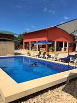 Uma piscina com espreguiçadeiras e guarda-sóis, um edifício vermelho com um pátio aberto e uma placa com a inscrição "RESIDENCE AINA".