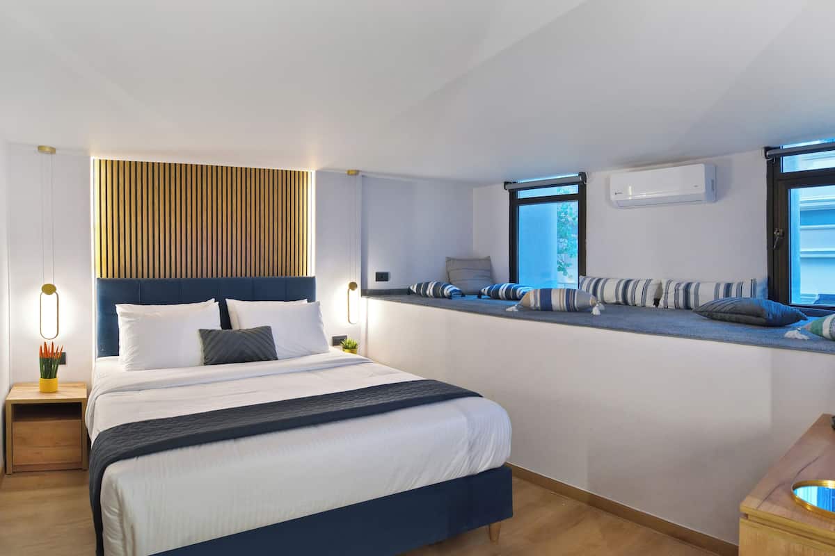 Quarto de hotel moderno com uma cama grande, um banco embutido com almofadas, uma mesa de cabeceira com abajur e uma janela com persianas.