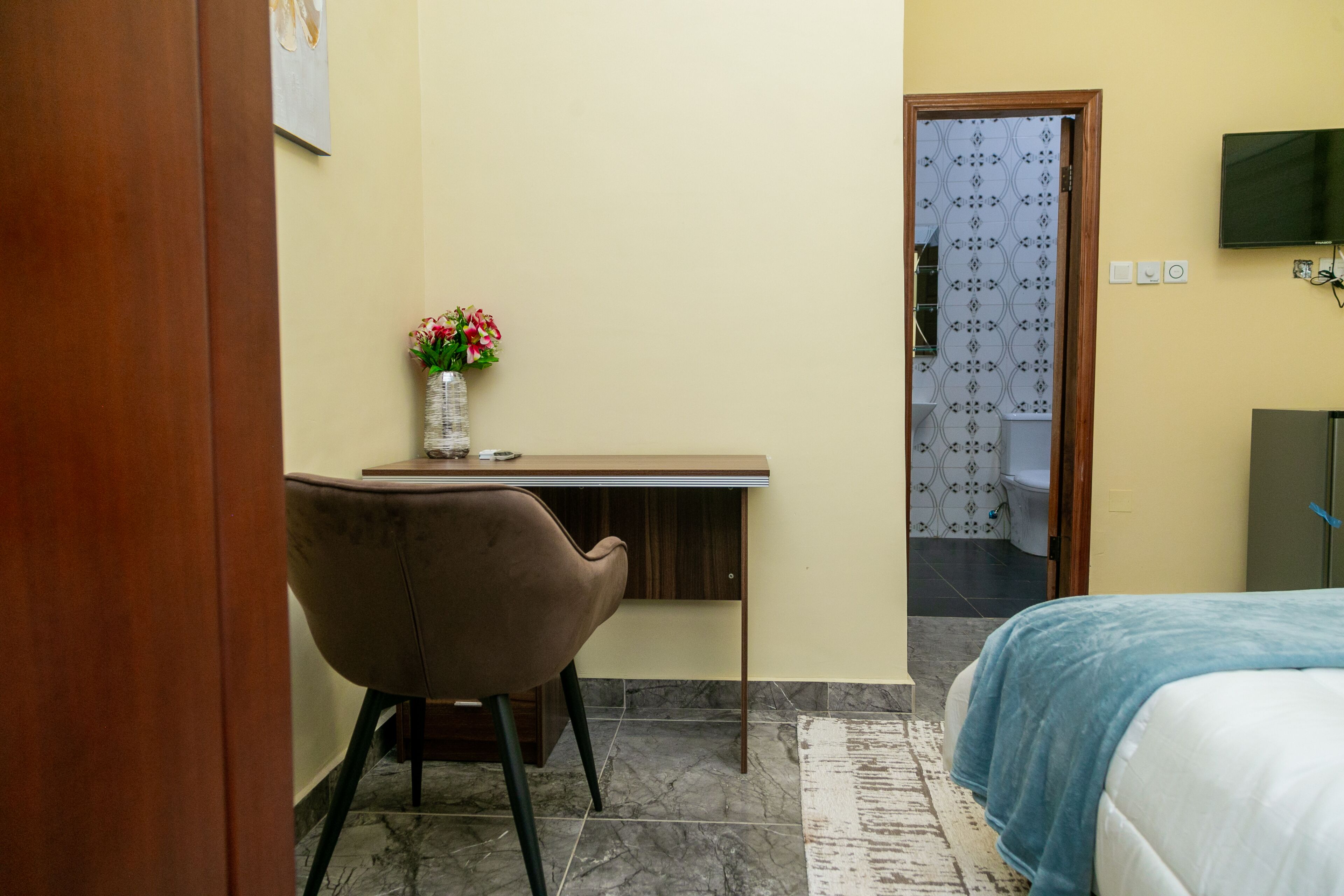 Quarto com cama, escrivaninha e cadeira, vaso com flores, e banheiro visível por uma porta.