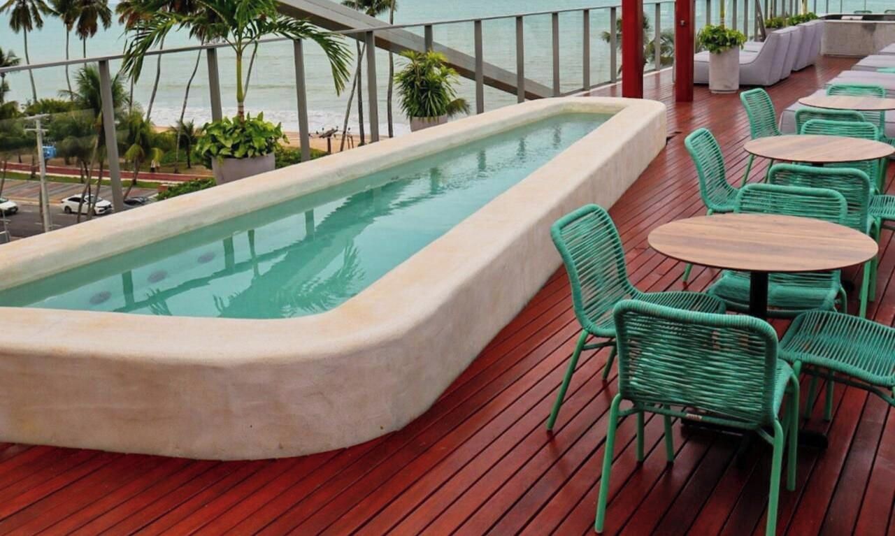 Área da piscina com deck de madeira, cadeiras verdes e vista para a praia.