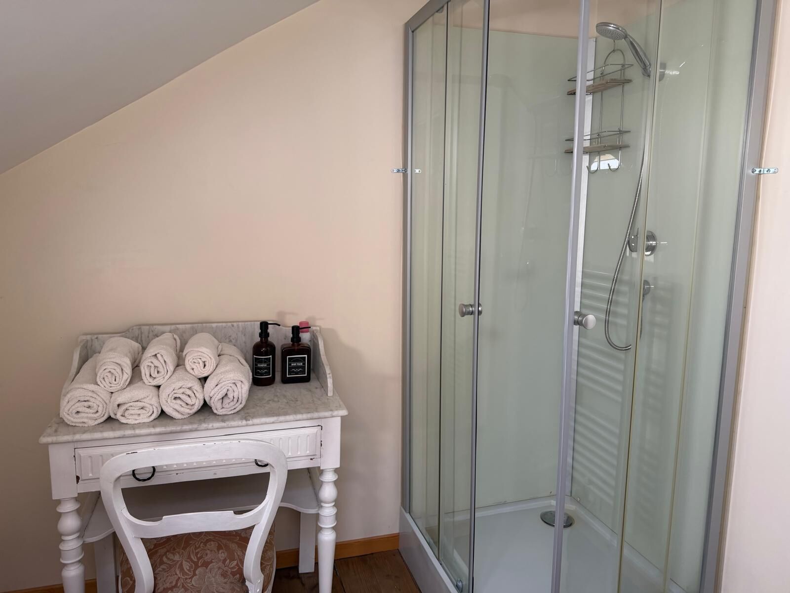 Um chuveiro com porta de vidro, uma mesa com toalhas enroladas e duas garrafas de shampoo.