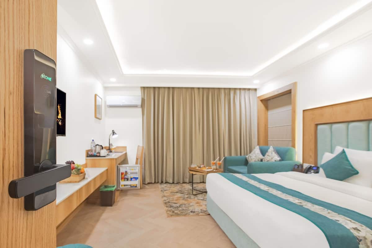 Quarto de hotel com cama, escrivaninha, televisão, poltrona, sofá e janela com cortinas.