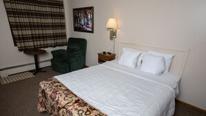 Quarto de hotel com uma cama grande, uma poltrona verde, uma mesinha com abajur e um quadro na parede.