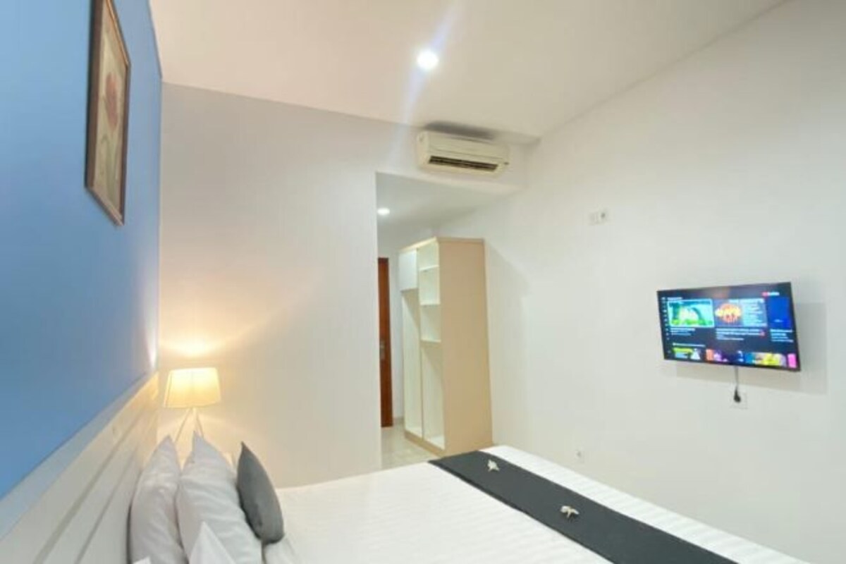Quarto de hotel com cama, televisão fixada na parede e abajur na mesinha de cabeceira.