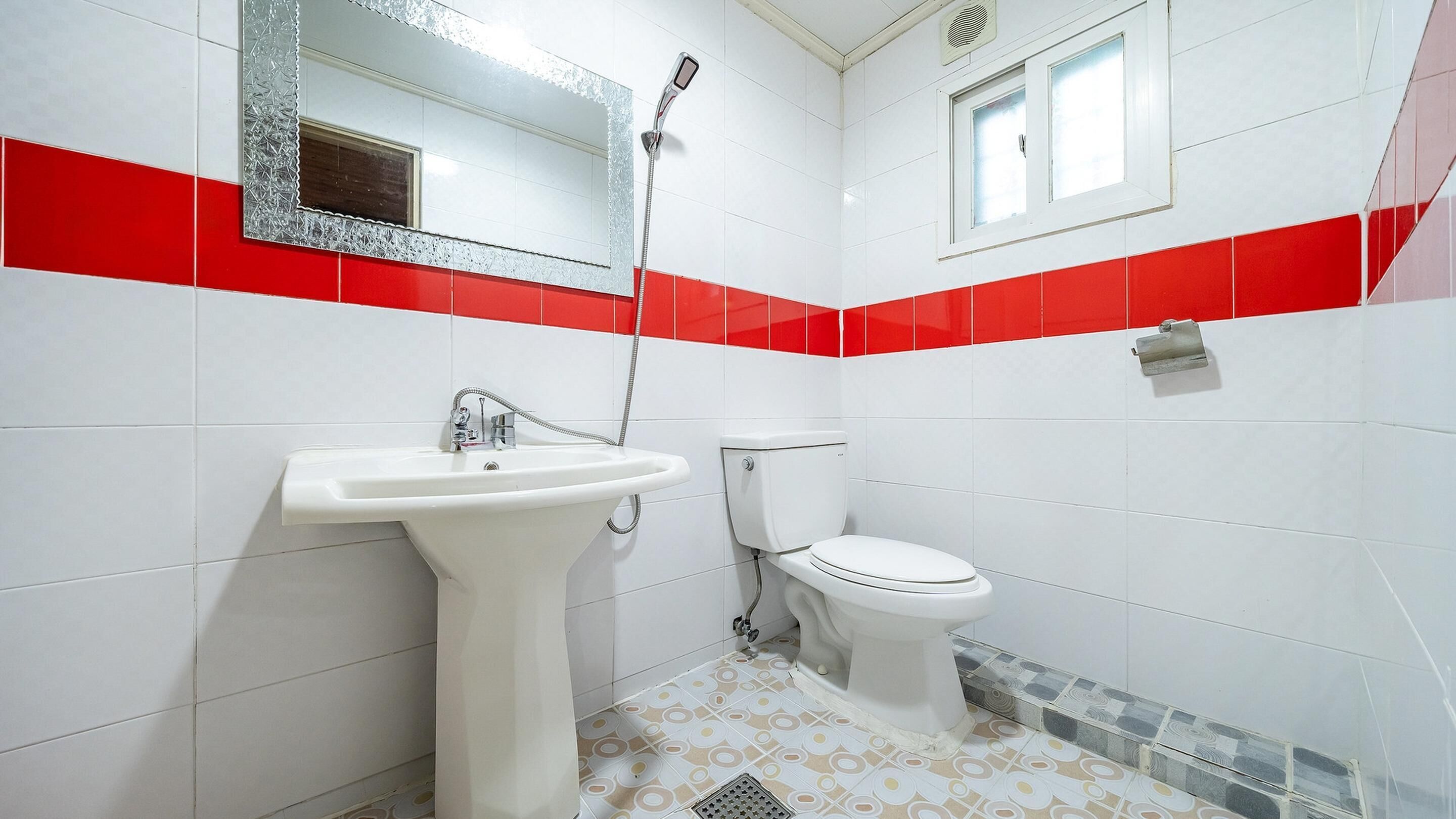 Casa de banho com lavatório, sanita e chuveiro brancos. As paredes estão revestidas a mosaicos vermelhos e brancos.