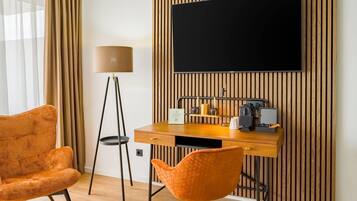Quarto de hotel moderno com escrivaninha de madeira, TV de tela plana, abajur e duas cadeiras laranja.