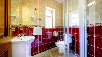 Casa de banho com lavatório branco, paredes com azulejos vermelhos, cabine de duche em vidro e uma janela.
