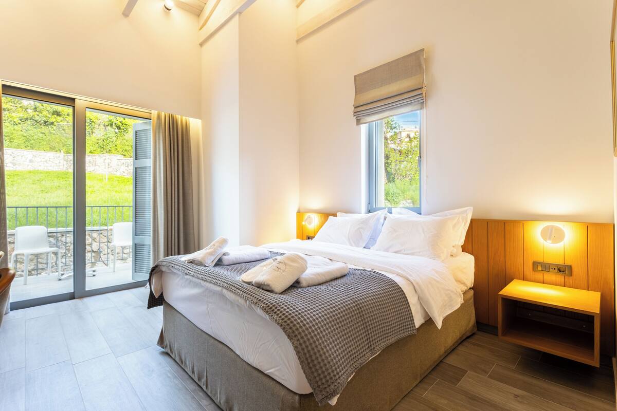 Quarto moderno com uma cama grande, mesa de cabeceira com abajur e porta de vidro deslizante para área externa.