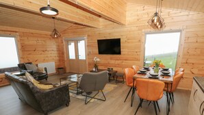 Uma sala de estar ampla com sofá, poltronas e uma mesa de jantar posta para quatro pessoas. Uma TV de tela plana está fixada na parede, e o ambiente possui paredes e pisos de madeira.