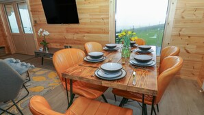 Uma sala de jantar com uma mesa de madeira posta para seis pessoas, cercada por cadeiras de couro laranja. Uma televisão de tela plana está fixada na parede acima da mesa.