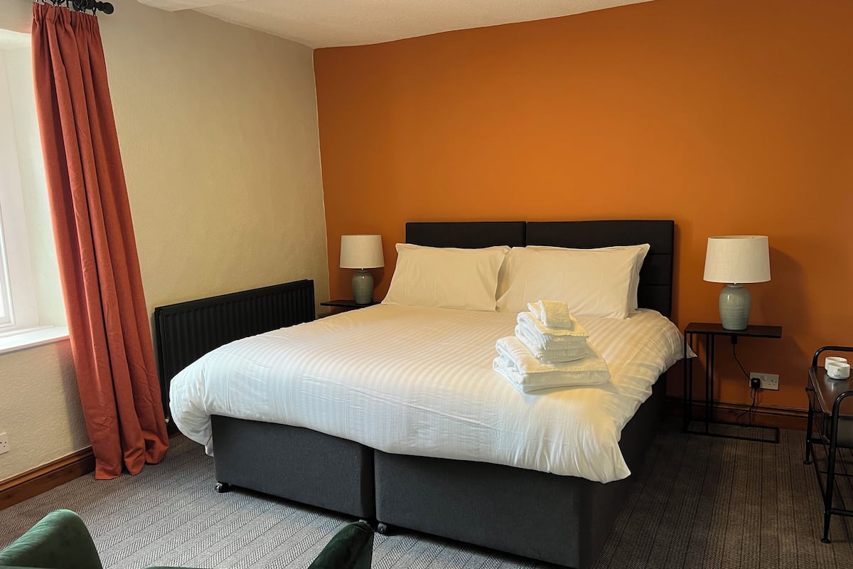 Quarto de hotel com cama, duas mesas de cabeceira com luminárias, uma poltrona verde e uma janela com cortinas.