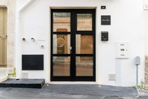 Entrada de um edifício moderno com porta de vidro emoldurada a preto, uma pequena área de estar ao ar livre e um letreiro com a inscrição 'Suite Mai'.