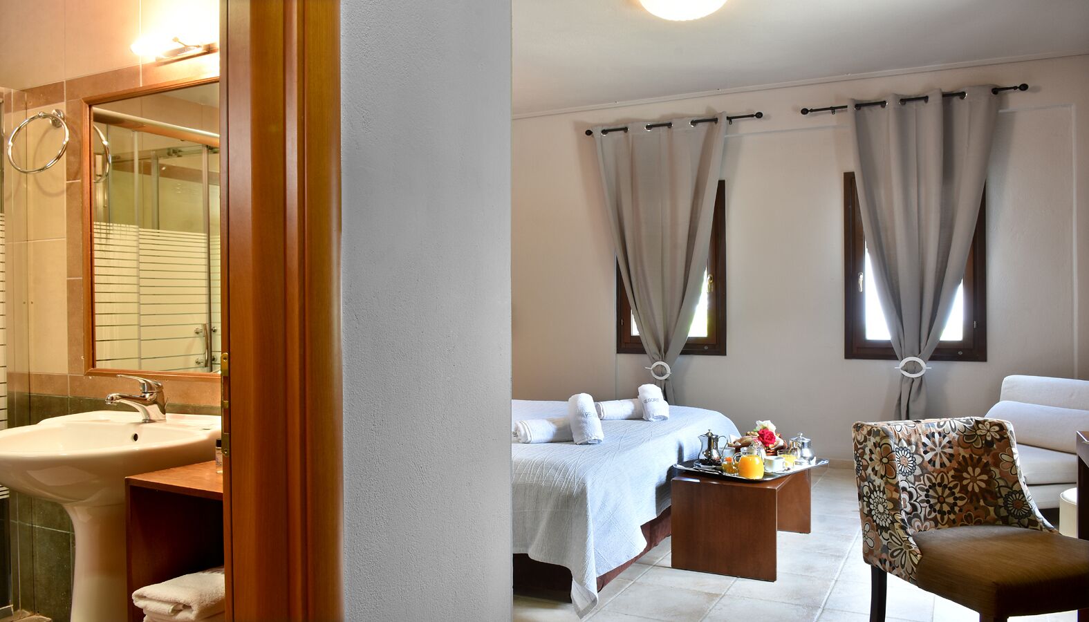 Quarto de hotel com banheiro, cama com toalhas, mesa de cabeceira com itens de café da manhã e uma cadeira.