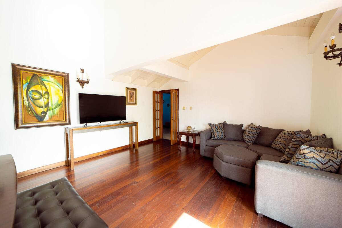 Sala de estar com piso de madeira, sofá cinza, banquinho preto, TV de tela plana em suporte de madeira e um quadro na parede.
