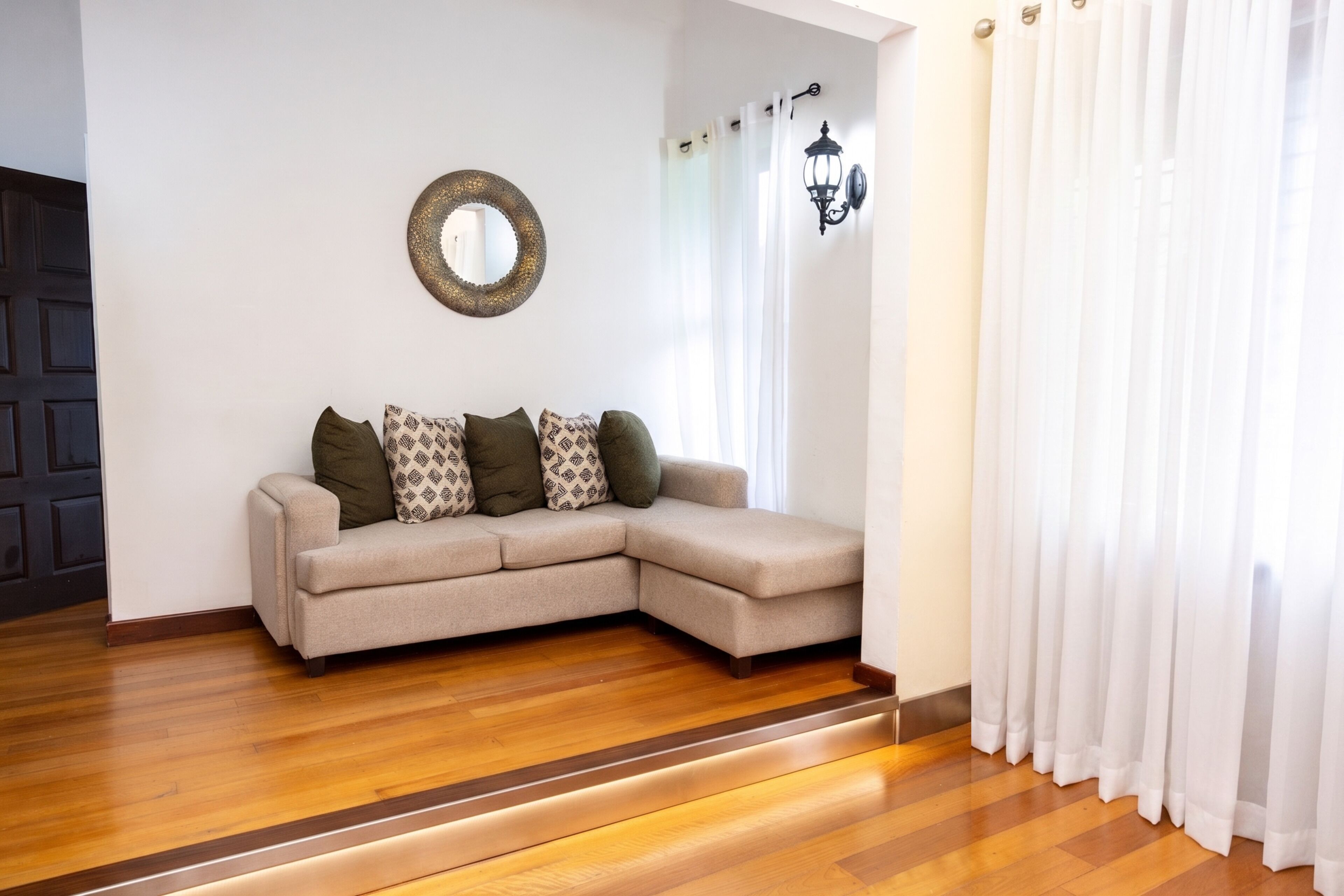 Uma sala de estar com um sofá modular bege, almofadas estampadas, um espelho redondo, um luminário fixado na parede e uma janela com cortinas brancas.