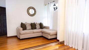 Uma sala de estar com um sofá modular bege, almofadas estampadas, um espelho redondo, um luminário fixado na parede e uma janela com cortinas brancas.