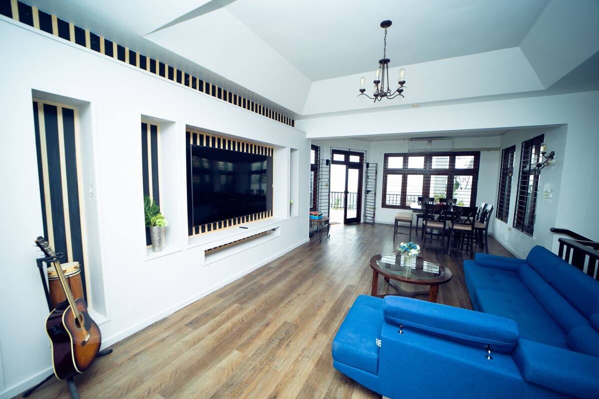 Uma sala de estar ampla, com uma televisão grande, um sofá azul e piso de madeira.