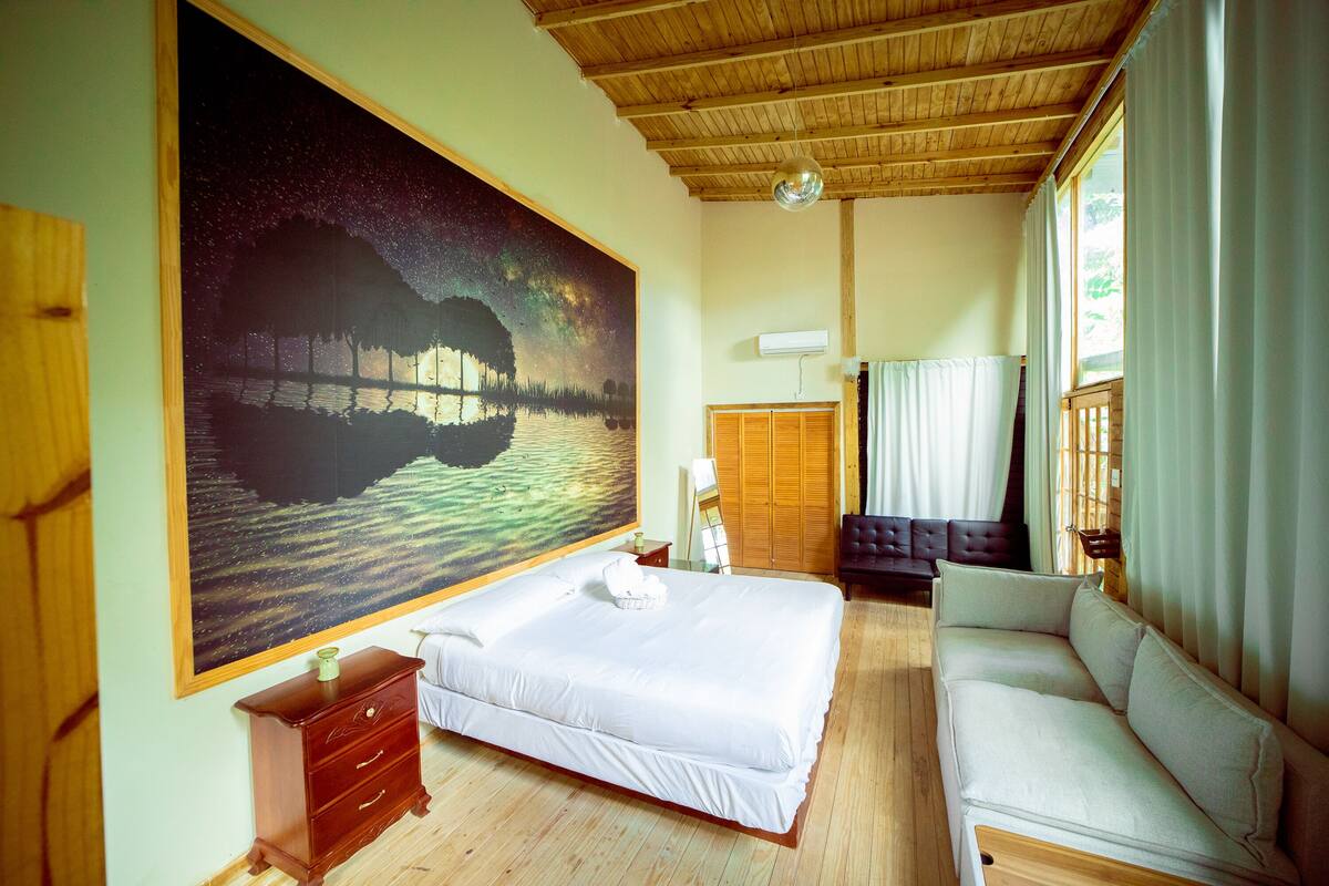 Quarto com uma cama grande, um sofá, cômoda de madeira, pintura representando uma cena noturna e janela com cortinas.
