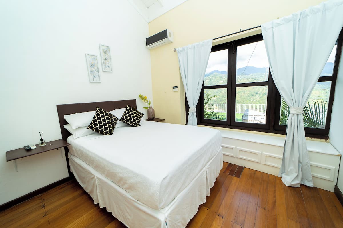 Cama bem arrumada com roupa de cama branca, mesa de cabeceira com abajur e janela com cortinas.