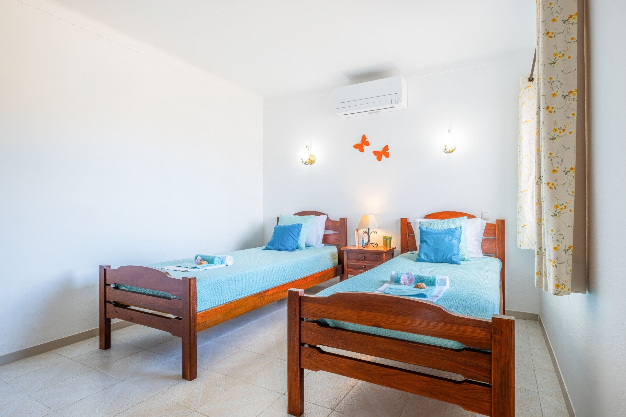 Quarto com duas camas de madeira, ar-condicionado fixado na parede e mesa de cabeceira com abajur.