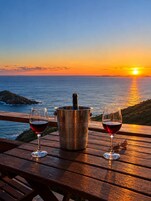 Vista do pôr do sol com mesa de madeira, dois copos de vinho tinto e um balde de metal com uma garrafa.