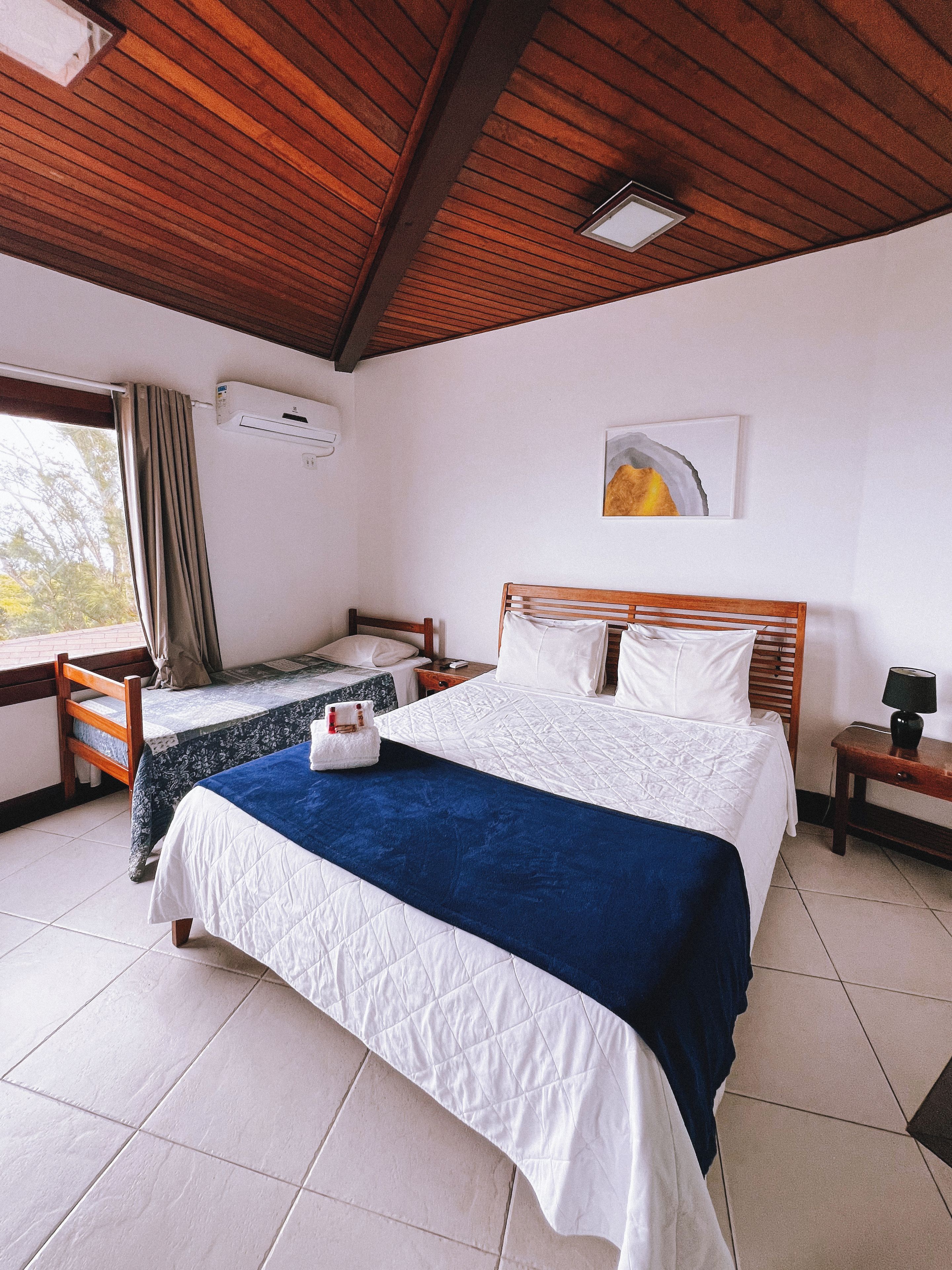 Quarto de hotel com uma cama grande, uma cama menor, janela com cortinas, um quadro na parede e um ventilador de teto.