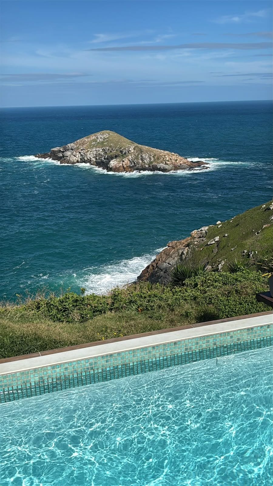 Piscina com água azul cristalina, rodeada por vegetação e com vista para um litoral rochoso e o oceano ao fundo.
