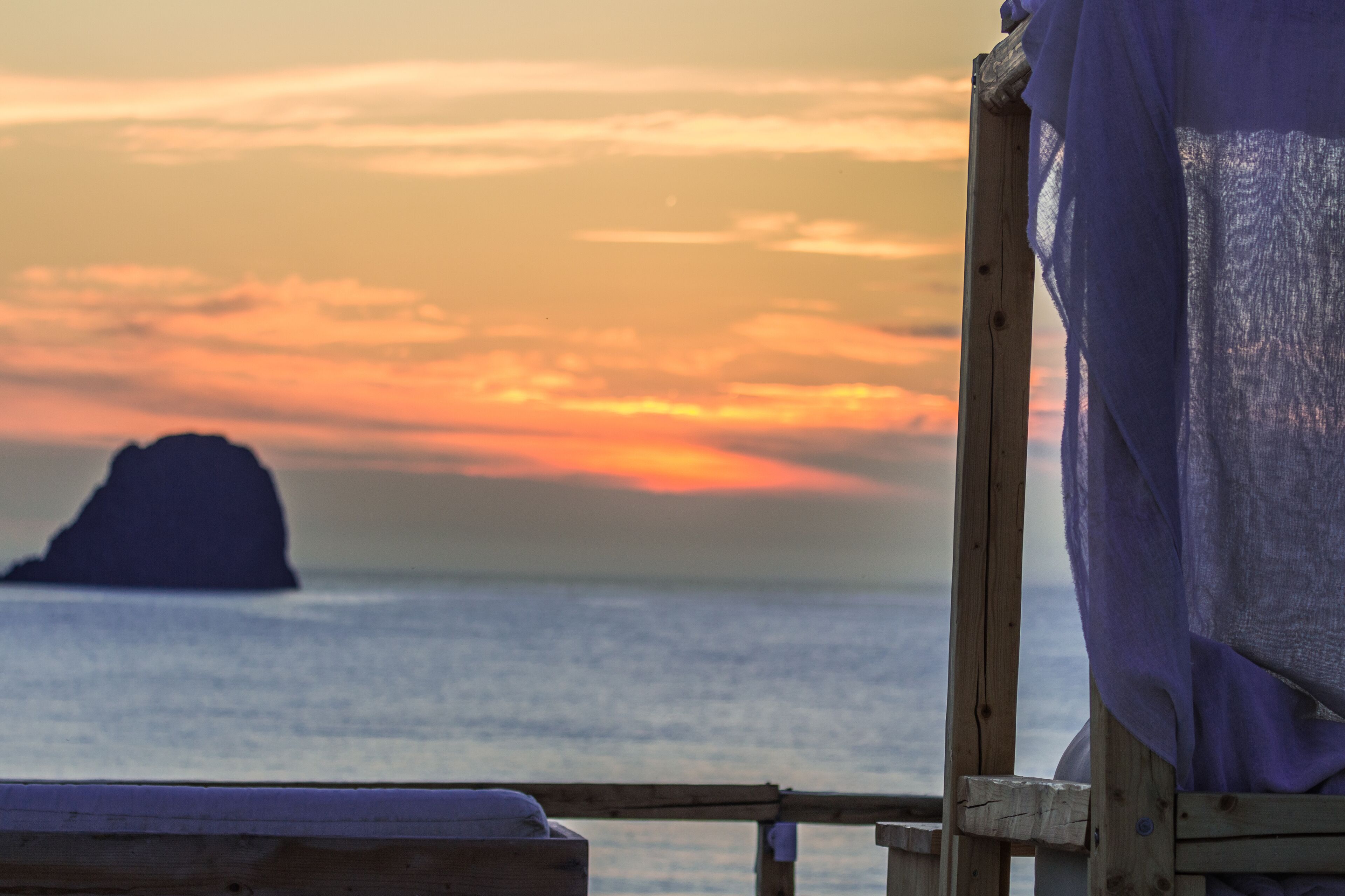 Um deck de madeira com vista para o oceano e uma ilha distante ao pôr do sol.