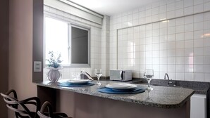 Cozinha com paredes revestidas de azulejos brancos, bancada de granito, micro-ondas, pia e janela com persianas.