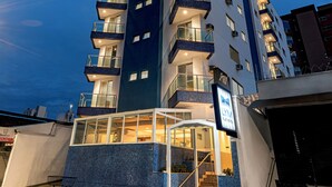 Um hotel de vários andares com design moderno, apresentando varandas em cada andar, uma entrada visível com escadas e uma placa indicando o nome do hotel.