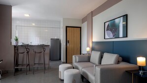 Uma sala de estar moderna com um sofá cinzento, dois pufes redondos e uma pequena mesa com uma luminária. Uma área de cozinha com um banco de bar e uma geladeira é visível ao fundo.