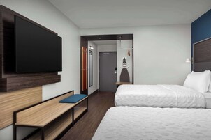 Quarto de hotel com duas camas, televisão presa na parede, um banco e porta para o banheiro.