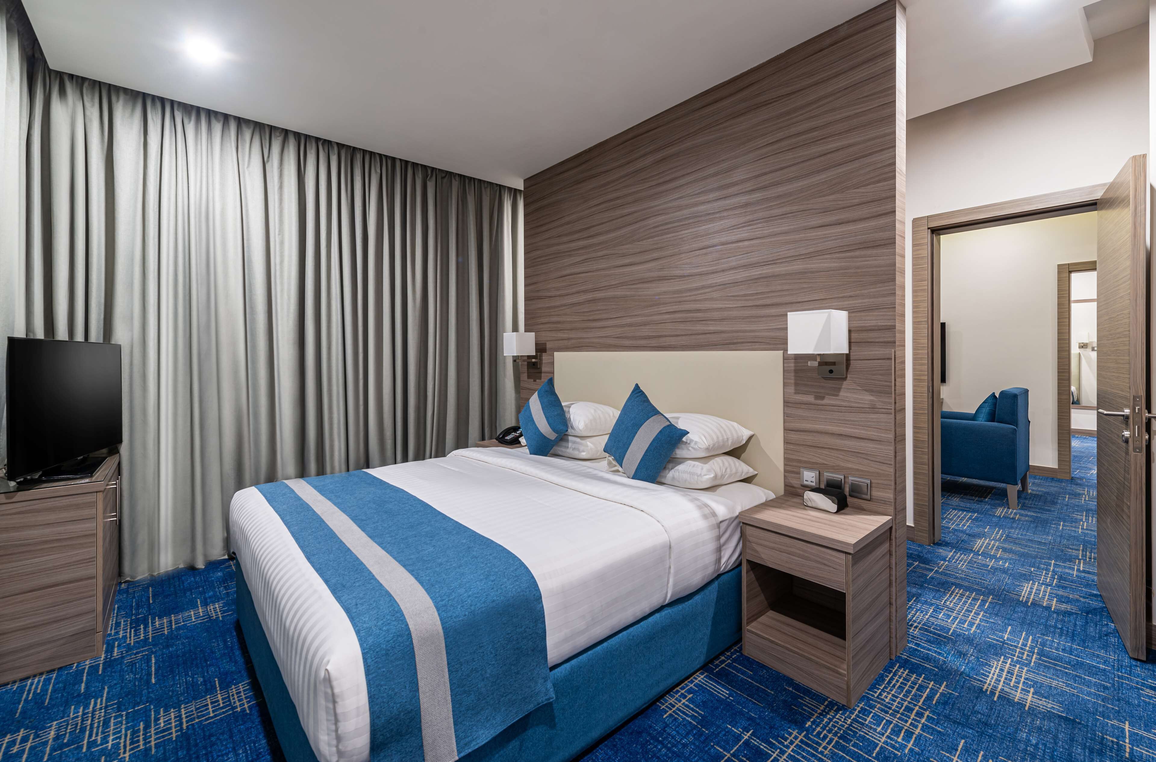 Quarto de hotel com uma cama grande, uma TV em um armário de madeira, uma escrivaninha com abajur e um tapete azul.