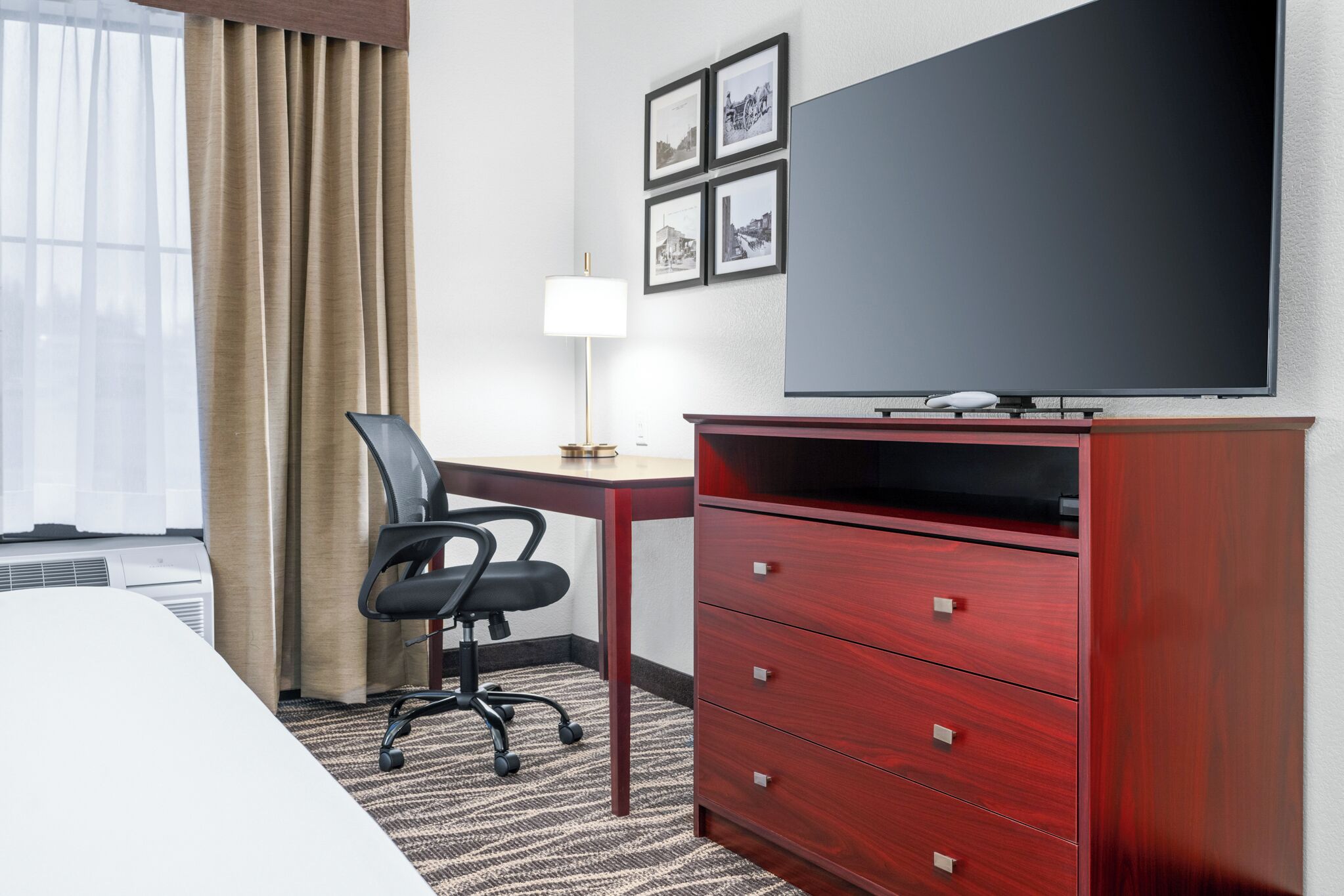 Quarto de hotel com uma cama grande, uma escrivaninha com cadeira, televisão, cômoda e janela com cortinas.