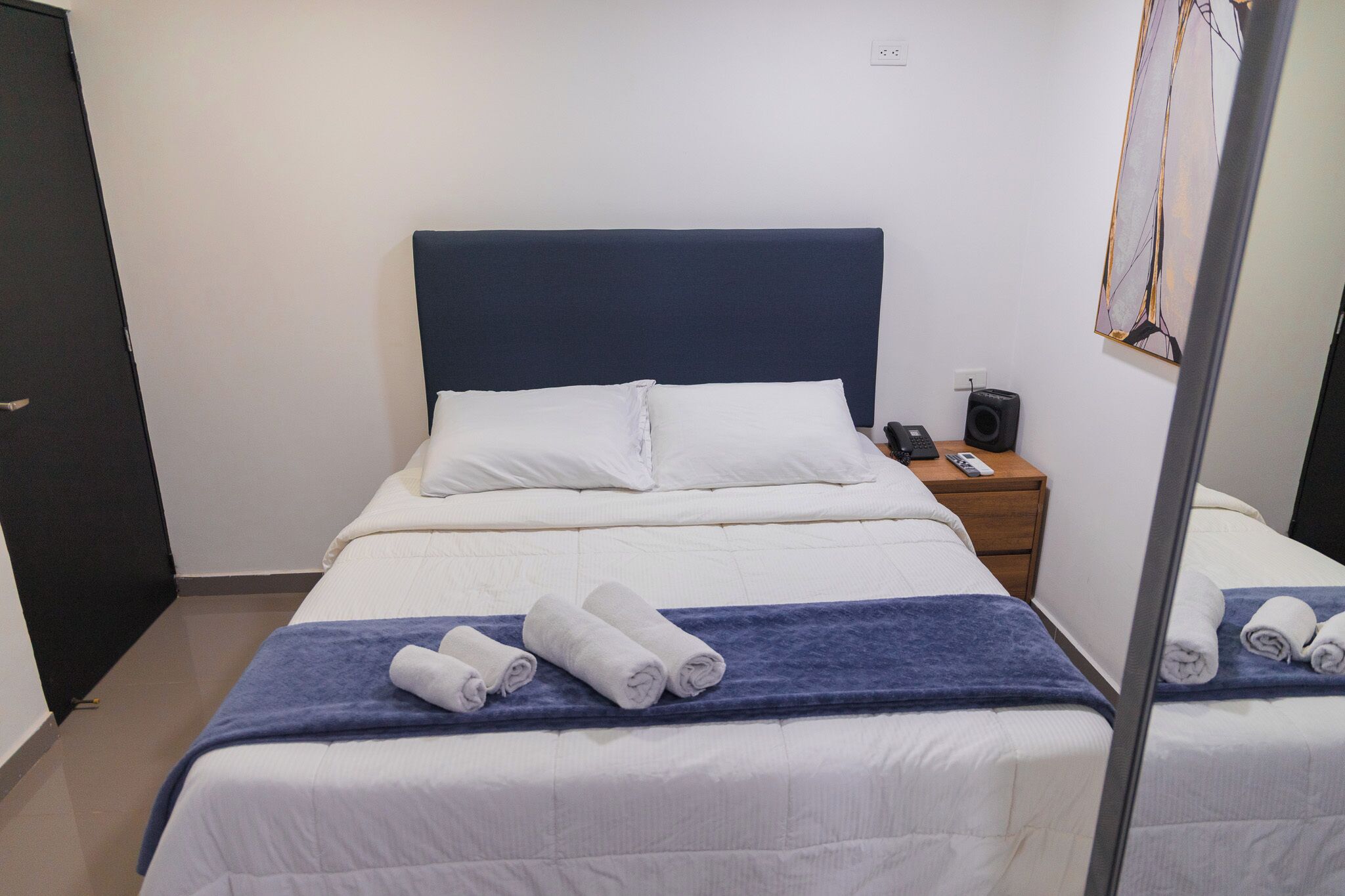 Quarto de hotel com cama, mesa de cabeceira, um alto-falante e um espelho.