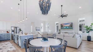 Uma cozinha ampla com eletrodomésticos modernos, uma sala de jantar com mesa e cadeiras redondas, e uma sala de estar com sofá e um quadro.